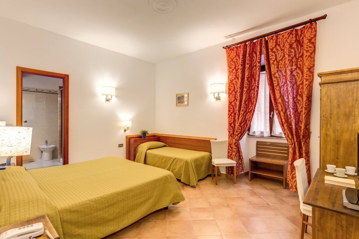 Quarto de hotel com duas camas, uma escrivaninha, uma cadeira, uma mesinha, um espelho, uma janela com cortinas e uma luminária na parede.