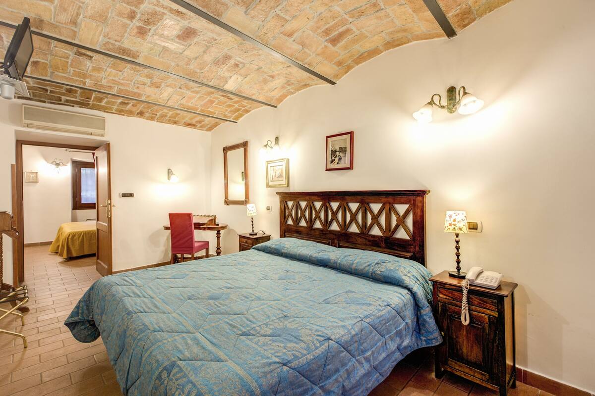 Quarto com uma cama grande, cabeceira de madeira, mesa de cabeceira com abajur, uma cadeira e uma pequena mesa com luminária.