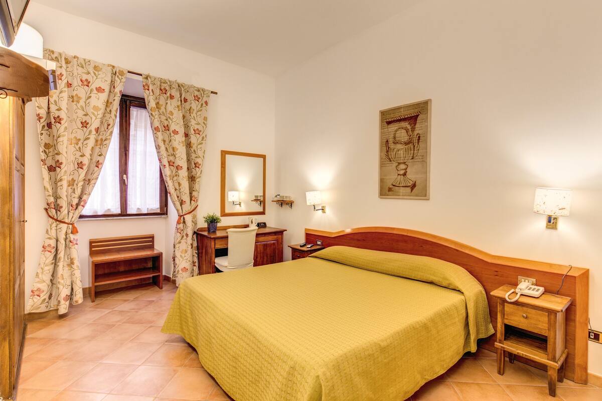 Quarto de hotel com cama, escrivaninha, cadeira, espelho, mesa de cabeceira com telefone e luminária na parede.