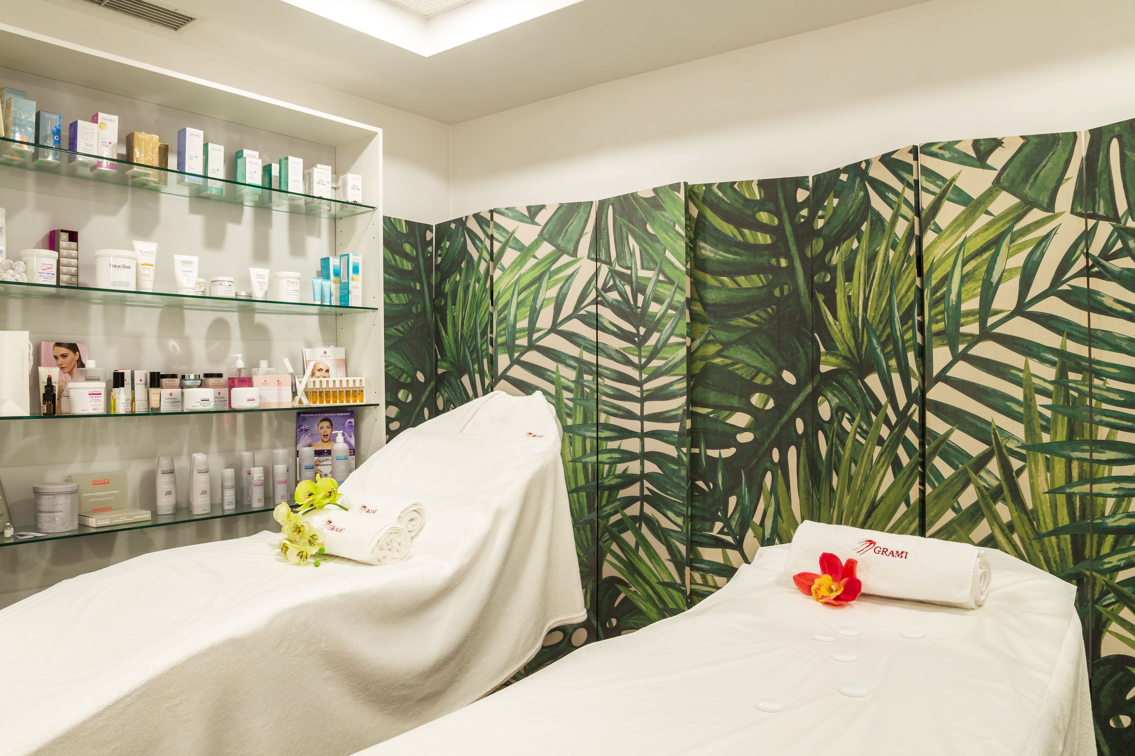 Uma sala de spa com duas camas cobertas por toalhas brancas, uma prateleira com diversos produtos de cuidados para a pele e um painel de parede com padrão de folha tropical.