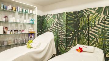 Uma sala de spa com duas camas cobertas por toalhas brancas, uma prateleira com diversos produtos de cuidados para a pele e um painel de parede com padrão de folha tropical.