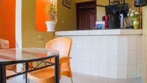 Uma área de refeições com uma mesa redonda e uma única cadeira laranja. Há uma planta em vaso na parede, e uma máquina de café está no balcão.