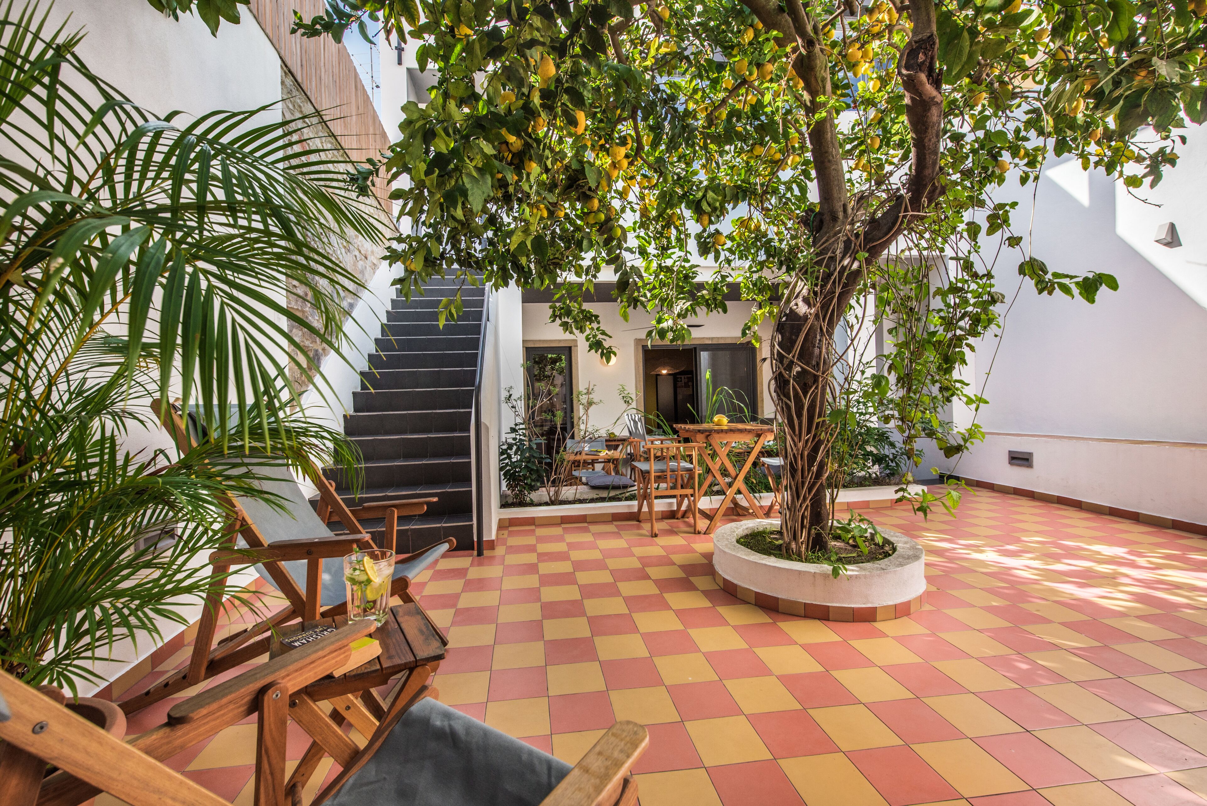 Um pátio com piso xadrez, móveis de madeira, uma árvore com frutos amarelos e uma escadaria.