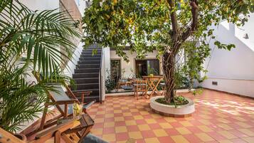 Um pátio com piso xadrez, móveis de madeira, uma árvore com frutos amarelos e uma escadaria.