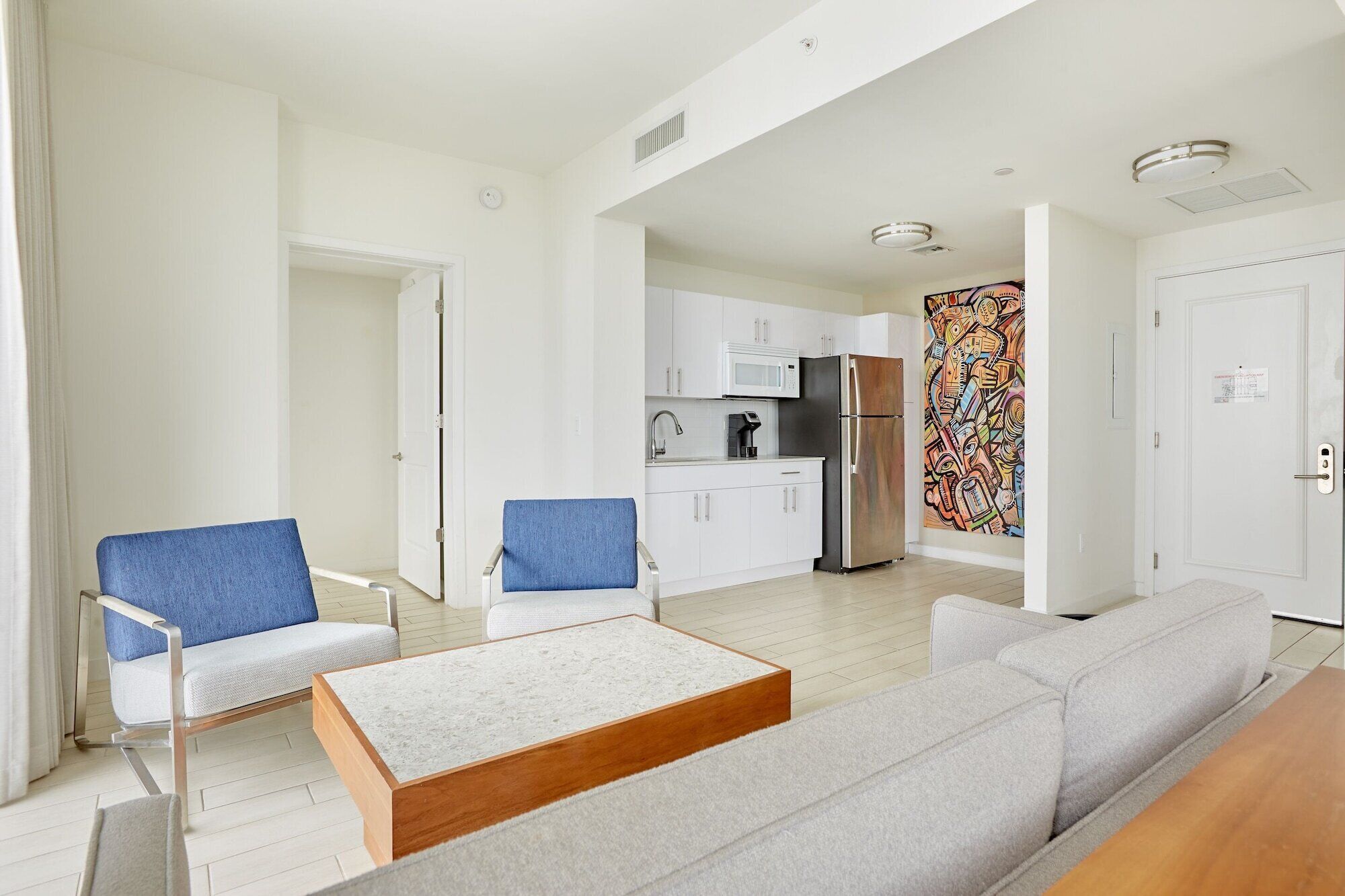 Uma sala de estar moderna com um sofá, duas poltronas, uma mesa de centro e uma área de cozinha com armários brancos, um micro-ondas e uma geladeira.