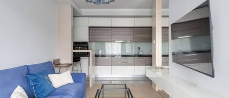 Uma sala de estar moderna com um sofá azul, uma mesa de centro de vidro e uma cozinha ao fundo.