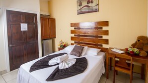 Uma cama bem arrumada com um cobertor branco e castanho, uma cabeceira de madeira, uma pequena mesa com um ursinho de pelúcia e flores, e uma porta com um aviso.