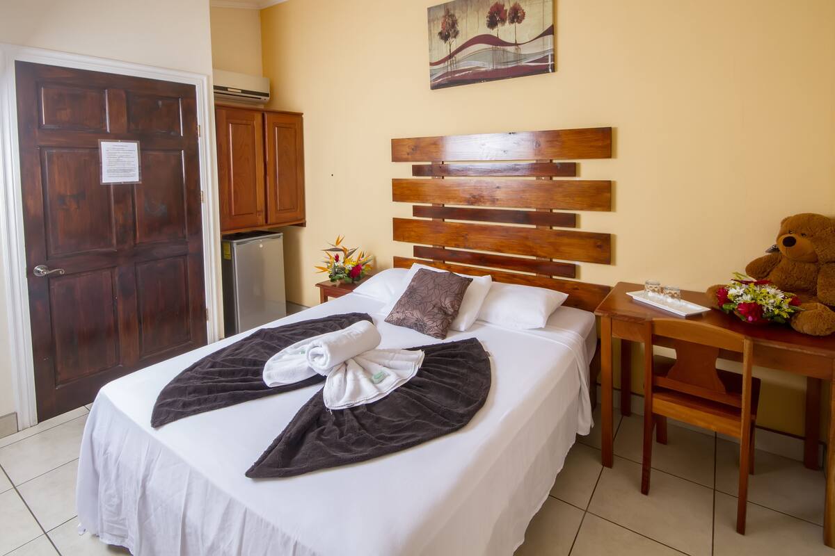 Uma cama bem arrumada com um cobertor branco e castanho, uma cabeceira de madeira, uma pequena mesa com um ursinho de pelúcia e flores, e uma porta com um aviso.