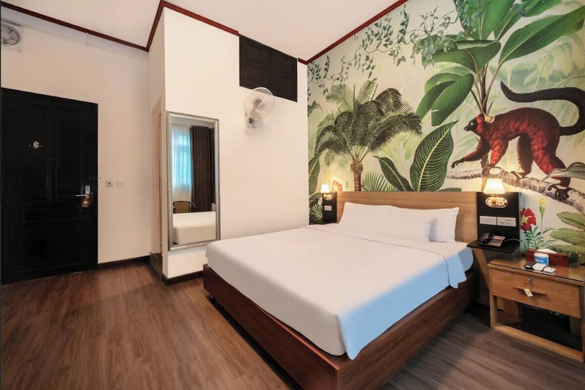 Quarto de hotel com uma cama grande, uma mesa de cabeceira, um espelho e um mural na parede com um macaco e folhagem.