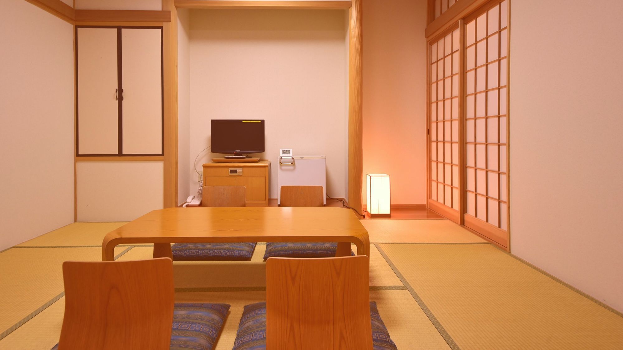 Um quarto japonês tradicional com tatames, uma mesa baixa com almofadas, uma televisão e um biombos shoji.