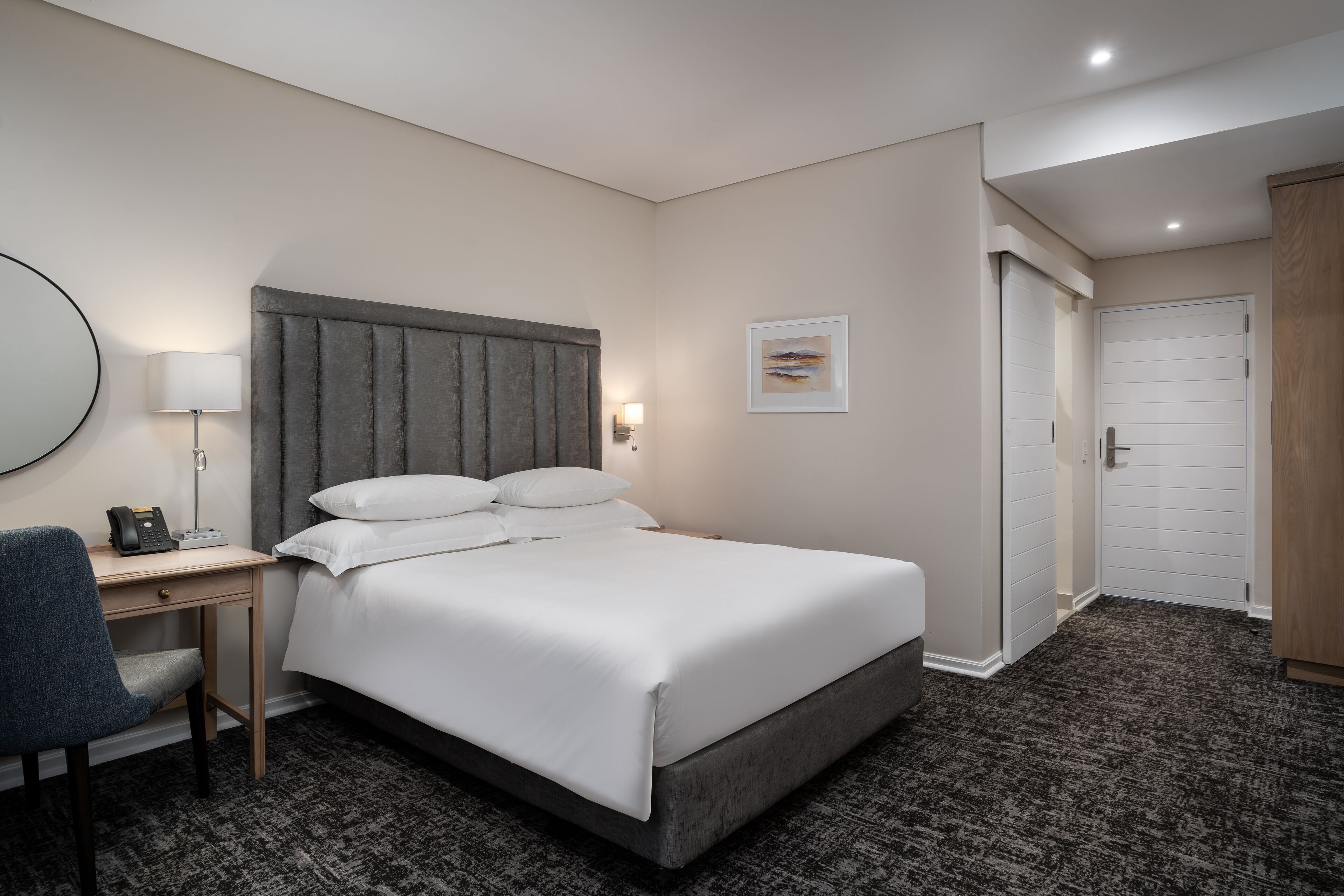 Quarto de hotel com cama, secretária, cadeira, luminária, quadro e guarda-roupa.