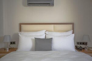 Uma cama bem arrumada com lençóis brancos e um travesseiro cinza, com duas mesas de cabeceira com luminárias e um telefone.