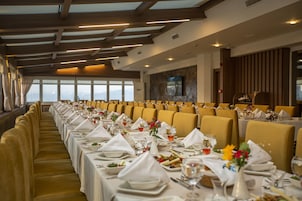 Uma longa mesa de jantar posta para uma refeição formal, com várias cadeiras, teto alto com vigas de madeira e vista para o exterior por grandes janelas.
