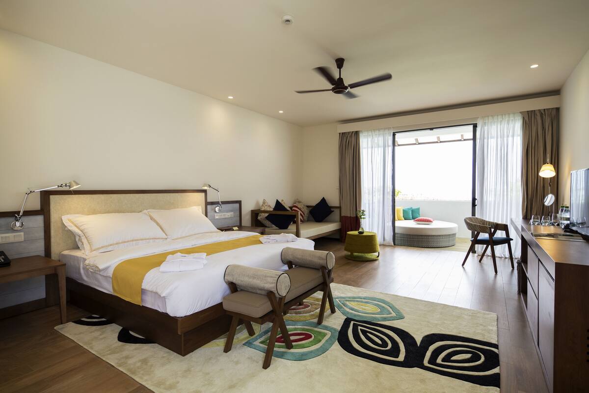 Quarto de hotel com cama, mesa de cabeceira, luminária, cadeira, escrivaninha e vista para o exterior.