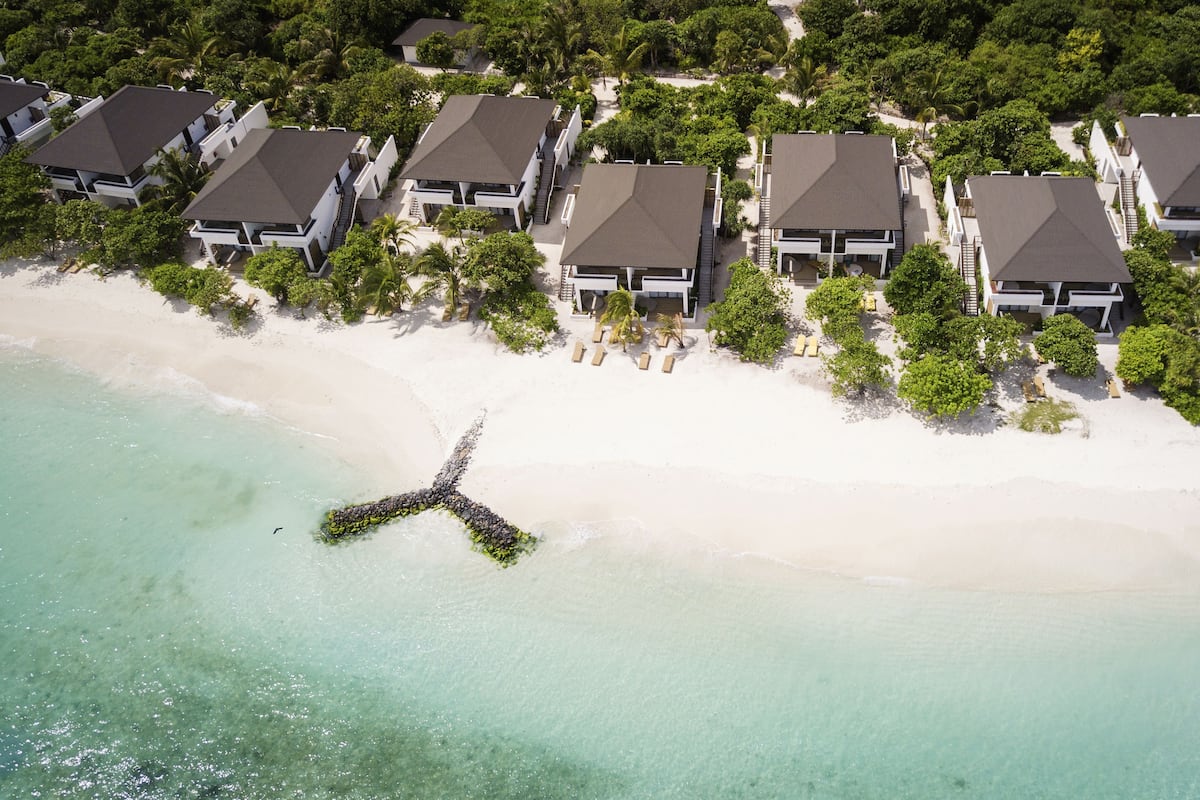 Vista aérea de um resort na praia com vários edifícios de dois andares, uma praia de areia e águas turquesa cristalinas.