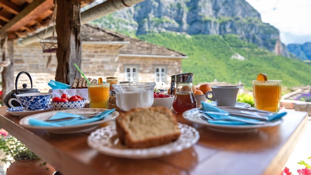 Mesa de café da manhã com diversos alimentos e vista para montanhas ao fundo.