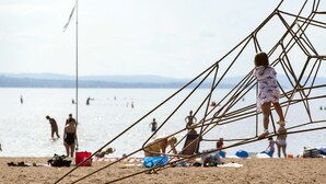 Uma praia com uma estrutura geométrica em primeiro plano e pessoas ao fundo.