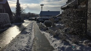 Rua residencial coberta de neve, com casas em ambos os lados, céu claro e um poste de luz visível.