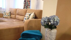 Uma sala de estar com um sofá bege, almofadas estampadas, um cobertor azul, um cesto de vime e um vaso com flores.