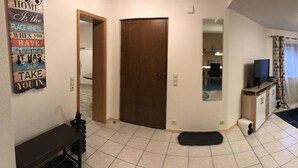 Uma sala de estar ampla com sofá, mesa de centro e televisão. Há uma área de jantar com mesa e cadeiras. O aposento possui uma grande janela com cortinas.