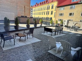 Uma área externa com mesas e cadeiras, plantas em vasos e vista para edifícios ao redor.