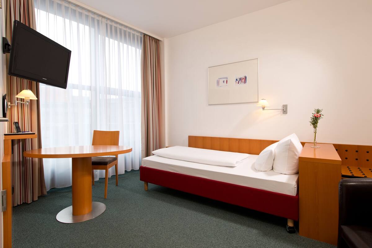 Quarto de hotel com cama, escrivaninha, cadeira, televisão e quadro na parede.