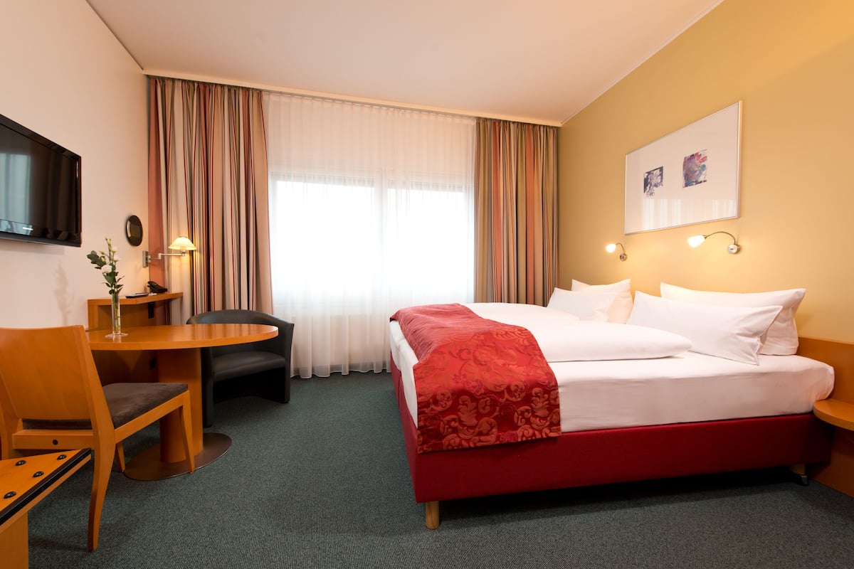 Quarto de hotel com cama, escrivaninha, cadeira, televisão e janela com cortinas.