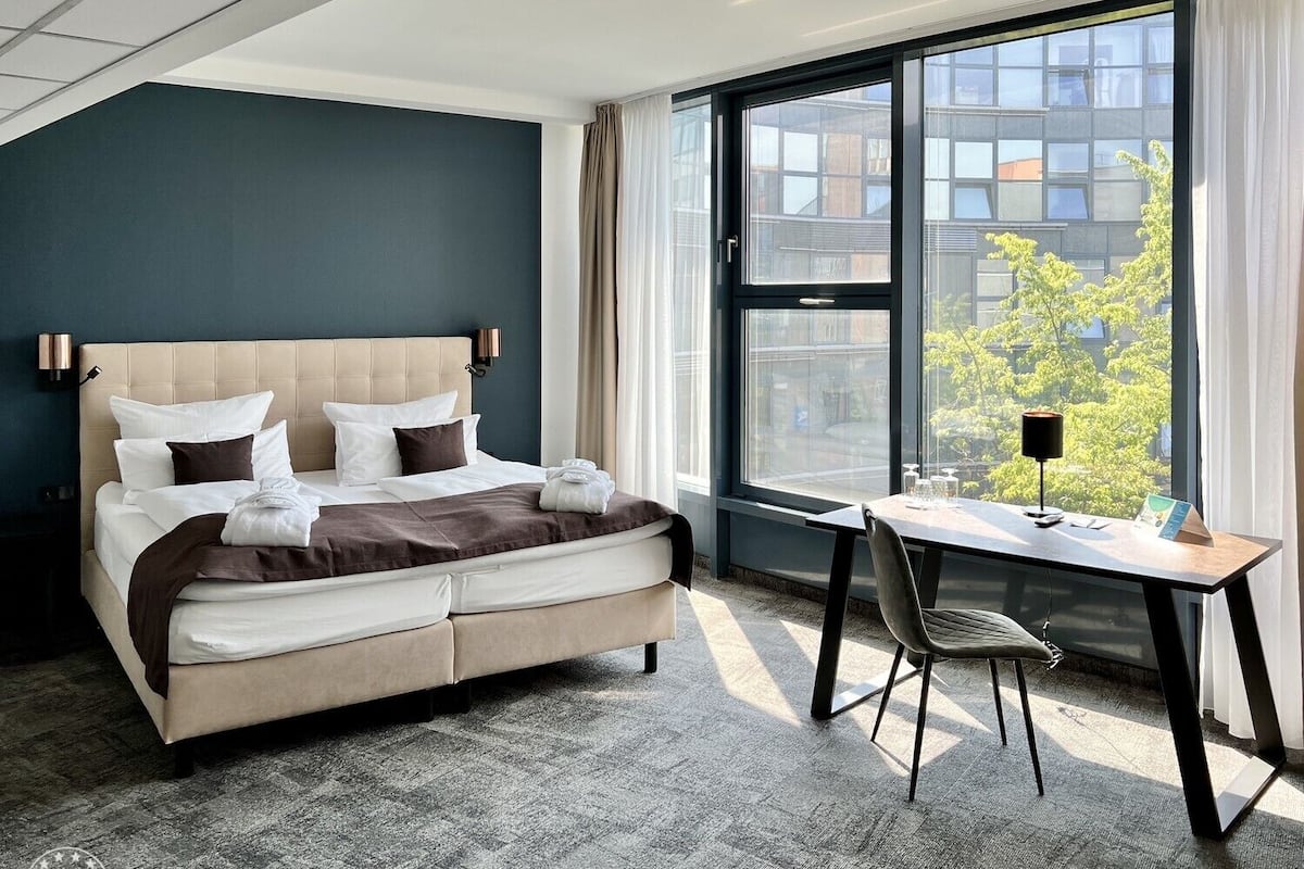 Quarto de hotel moderno com uma cama grande, uma escrivaninha e vista para a cidade através de amplas janelas.