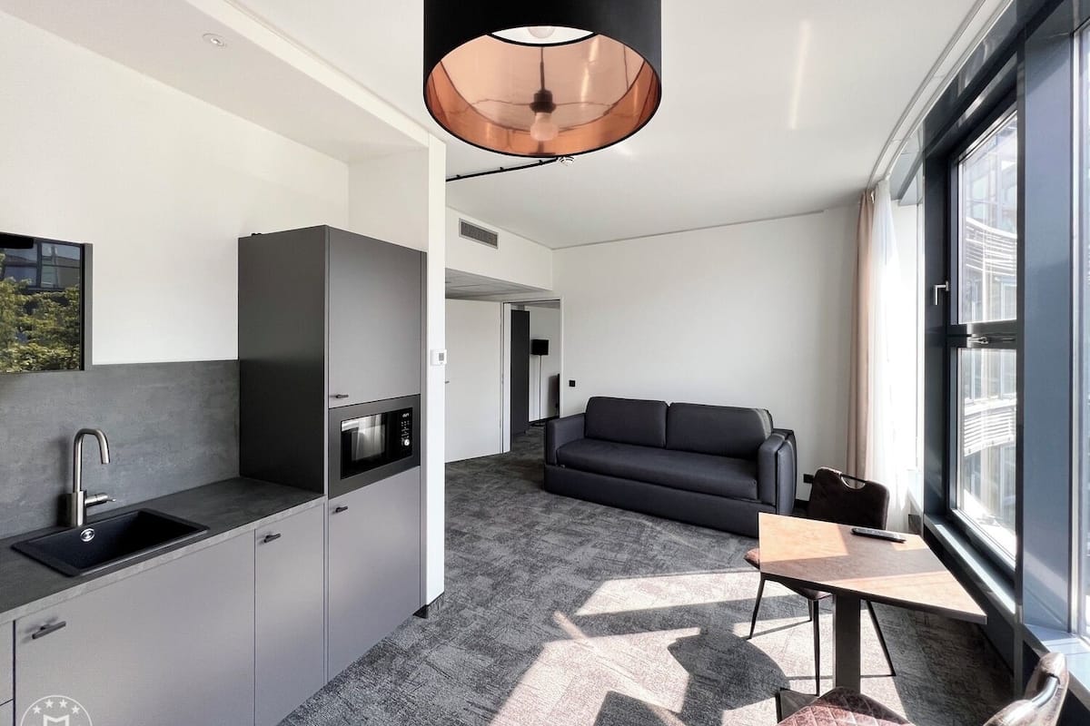 Cozinha moderna com micro-ondas embutido, pia e luminária pendente. Na sala de estar, um sofá escuro e uma mesa pequena com cadeiras, além de uma grande janela que proporciona luz natural.