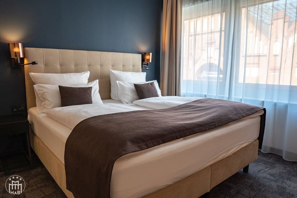 Quarto de hotel com uma cama grande, cabeceira, duas mesas de cabeceira, uma janela com cortinas e vista para edifícios.