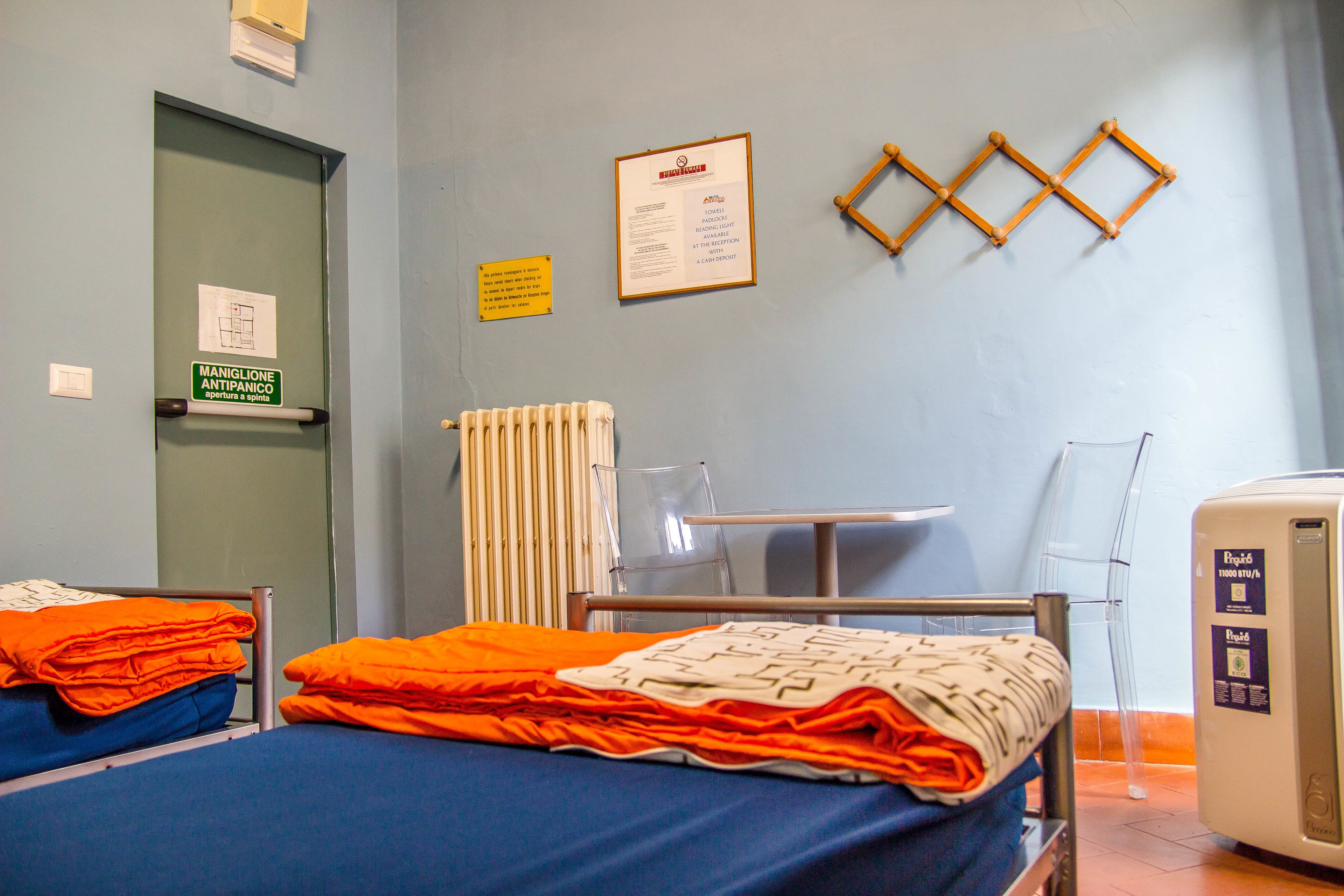 Quarto de hotel com duas camas, um radiador, uma escrivaninha e uma cadeira. Há placas nas paredes e um mural de avisos.