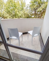 Uma varanda com duas cadeiras de plástico branco e uma mesa, com vista para um jardim com árvores.