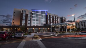Um hotel moderno com fachada de vidro, uma placa chamativa e um estacionamento com rastros de luz de veículos em movimento.