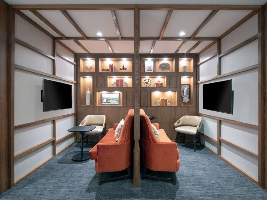 Quarto com dois sofás laranja, uma TV à esquerda e outra à direita. Há prateleiras com diversos objetos e o chão é coberto por carpete.