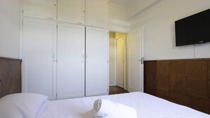 Quarto de hotel com cama, guarda-roupa branco, cabeceira de madeira e televisão fixada na parede.