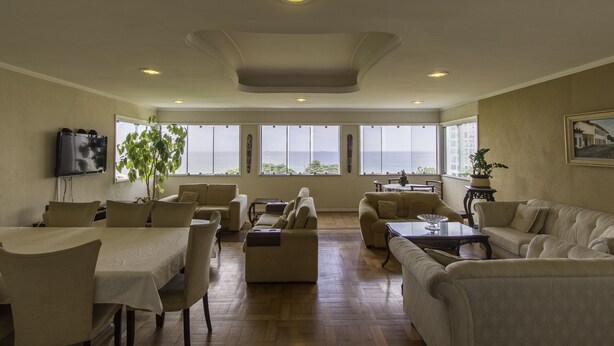 Ampla sala de estar com grande janela com vista para o mar, equipada com sofás, mesa de jantar e televisão na parede.