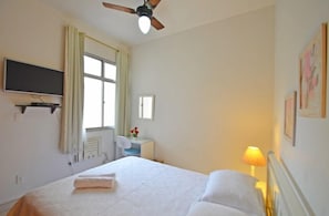Quarto de hotel com cama, escrivaninha, cadeira, TV fixada na parede, ventilador de teto e janela com cortinas.