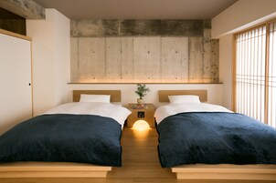 Quarto moderno com duas camas, teto de concreto, cabeceiras de madeira e uma mesinha de cabeceira com uma planta.