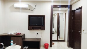 Quarto de hotel com televisão de tela plana na parede, uma pequena mesa com uma cadeira, escrivaninha com telefone e garrafa de água, um balde vermelho e porta que dá para outro quarto.