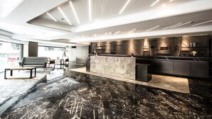Um lobby moderno com piso de mármore, um balcão de recepção, uma área de estar com sofá e cadeiras, e um espaço de cozinha com bancada e eletrodomésticos.