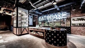 Cozinha moderna com um letreiro de neon que diz 'EAT@EASE', prateleiras com utensílios de cozinha e uma ilha central com uma mesa preta e cruzes brancas.