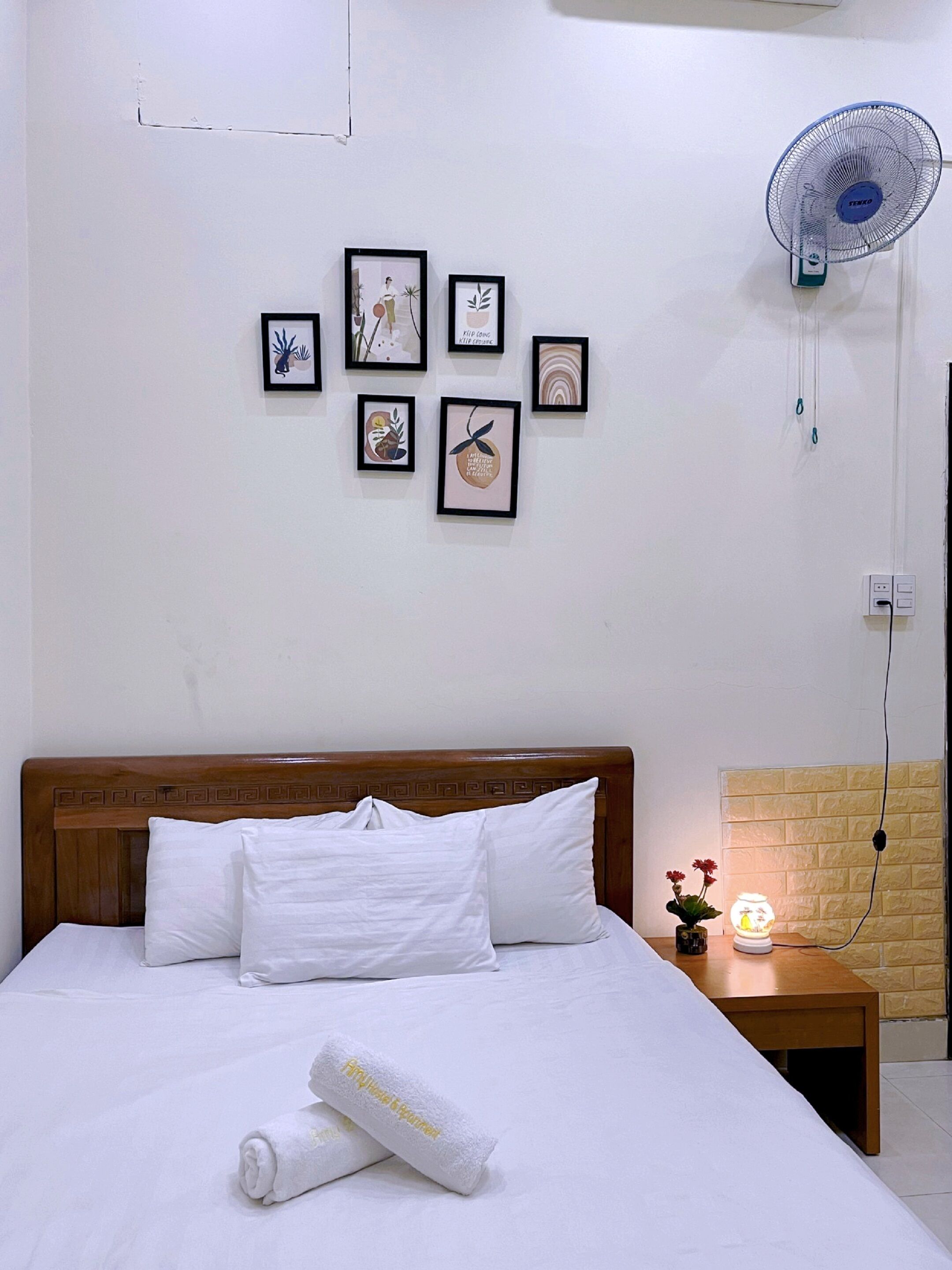 Uma cama bem arrumada com lençóis brancos e uma toalha enrolada, uma pequena mesa de cabeceira com abajur e um ventilador fixado na parede.