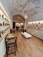 Um quarto amplo com piso de madeira, uma mesa comprida de madeira e várias cadeiras. Prateleiras nas paredes exibem diversos frascos e potes.