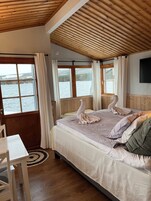 Quarto com teto de madeira, cama com edredom branco e cinza, dois travesseiros em formato de cisne, uma cadeira branca e vista para um lago através de uma janela.