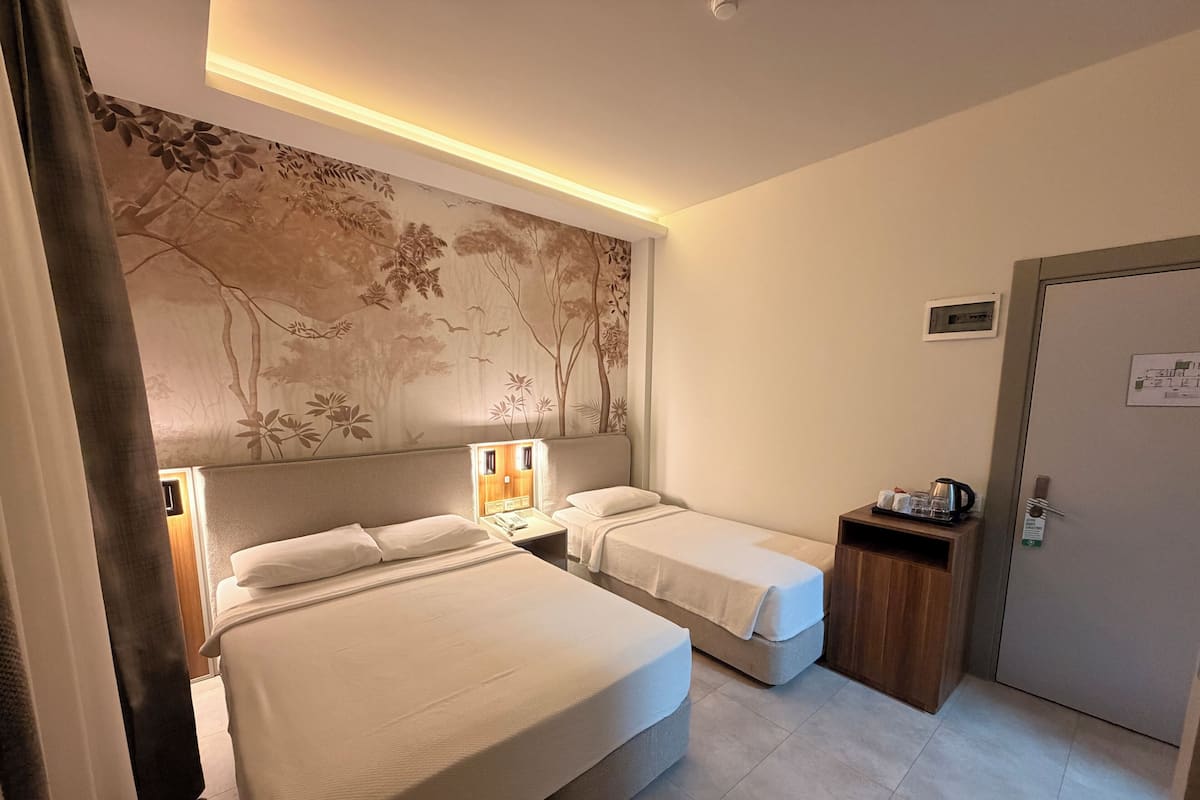 Quarto de hotel com duas camas, um grande mural na parede, luminárias de cabeceira, uma mesinha com um chaleira e uma porta com uma placa.