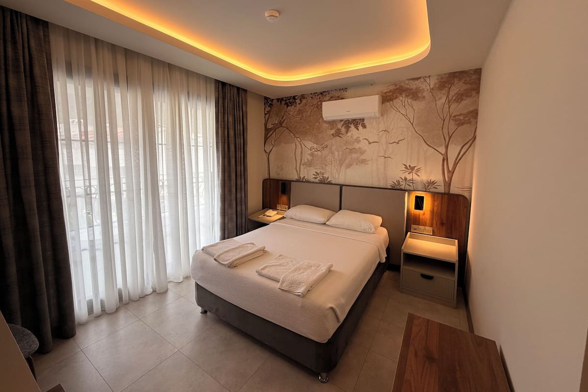 Quarto de hotel moderno com uma cama grande, mesas de cabeceira, ar condicionado e vista através de cortinas leves.