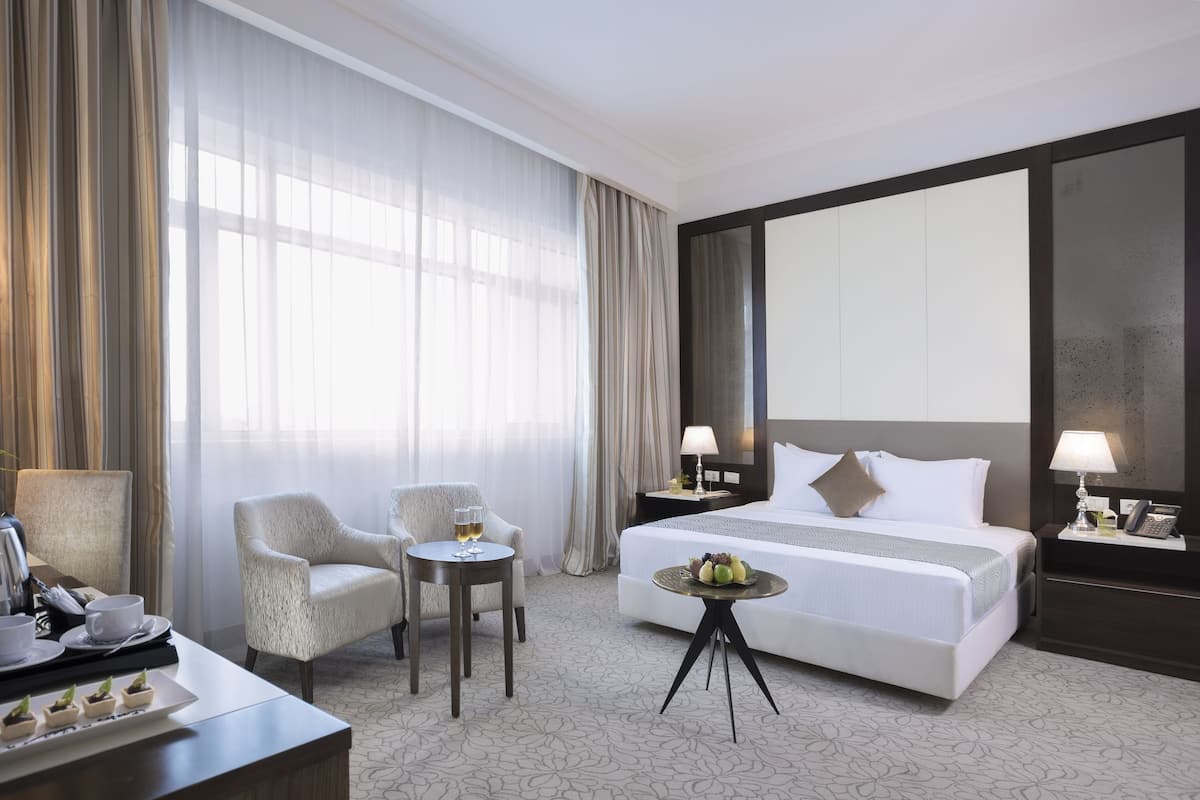 Quarto de hotel com uma cama grande, duas poltronas, uma mesinha redonda com uma bebida, uma cafeteira e uma bandeja com lanches.