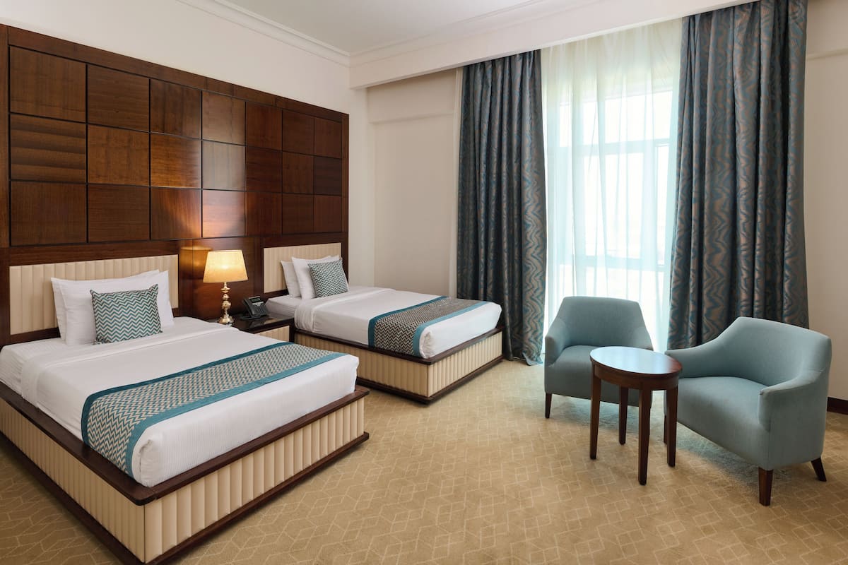 Quarto de hotel com duas camas, uma mesa pequena, uma cadeira e uma janela grande com cortinas.