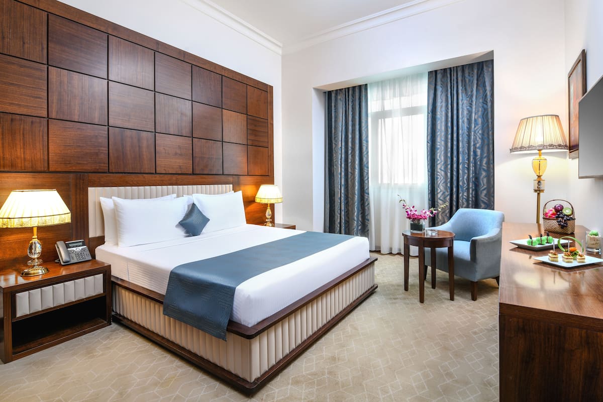 Quarto de hotel com uma cama grande, duas mesas de cabeceira com abajures, uma mesinha com um vaso de flores, uma cadeira e uma escrivaninha com uma cesta de frutas e um copo de água.