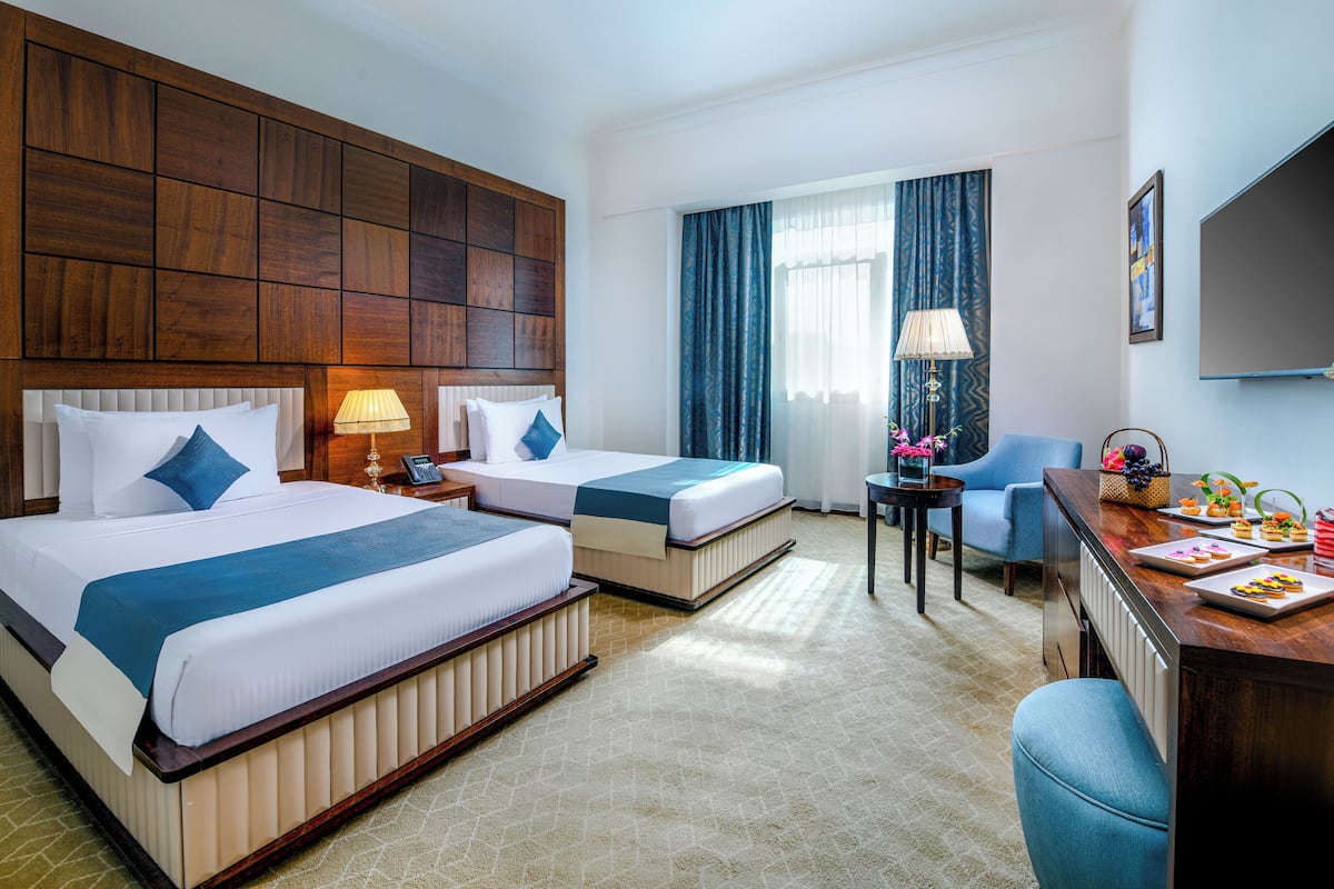 Quarto de hotel com duas camas, uma escrivaninha, uma cadeira, televisão e janela com cortinas.